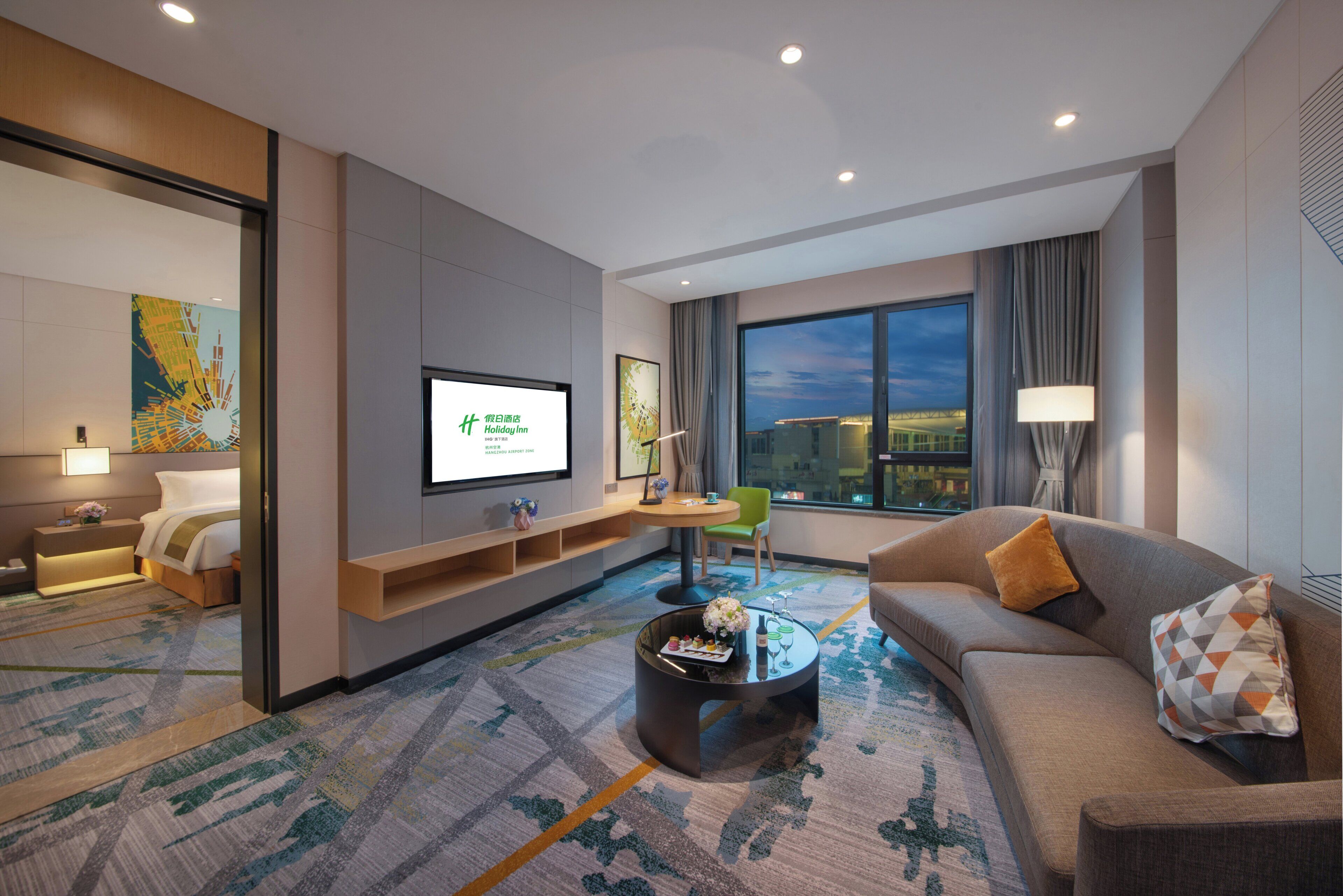 Quarto de hotel com cama, sofá, mesa de centro, televisão e uma grande janela com vista para a cidade.