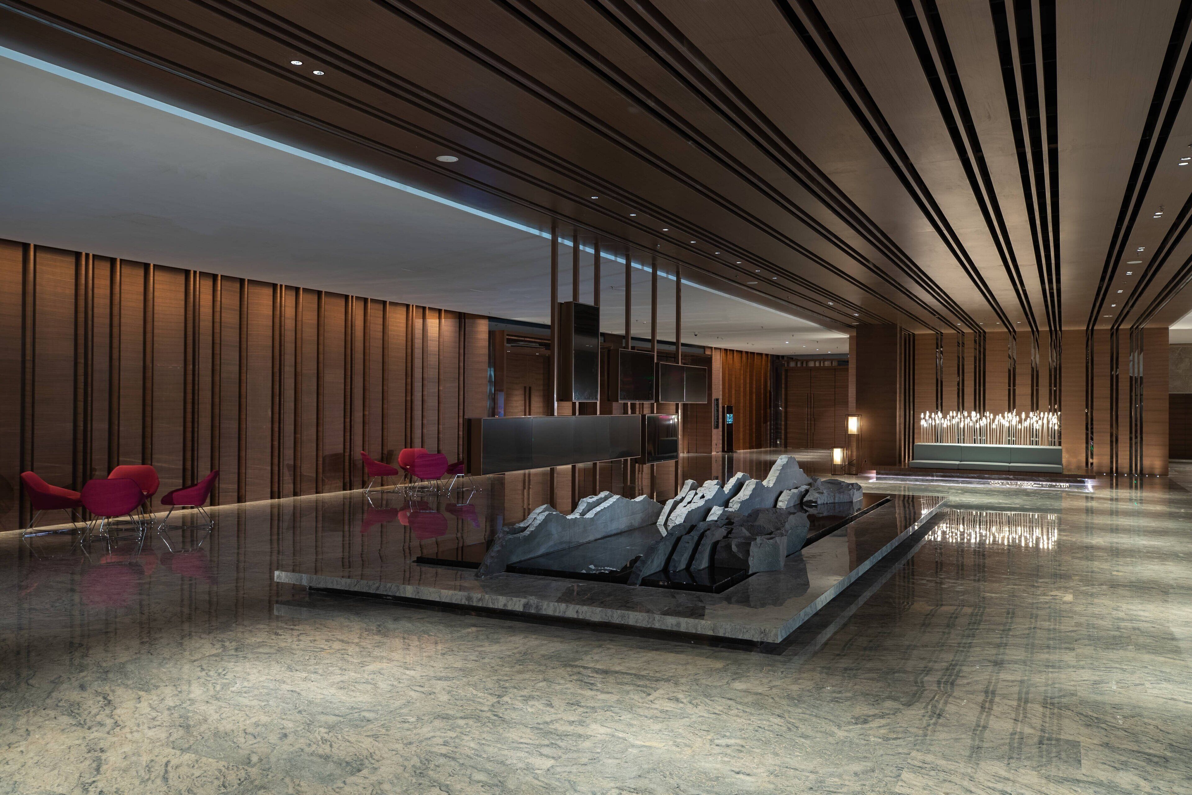 Um lobby moderno com balcão de recepção, cadeiras vermelhas, uma escultura de pedra e um candelabro.