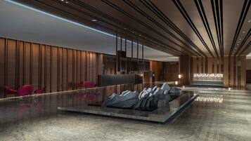 Um lobby moderno com balcão de recepção, cadeiras vermelhas, uma escultura de pedra e um candelabro.