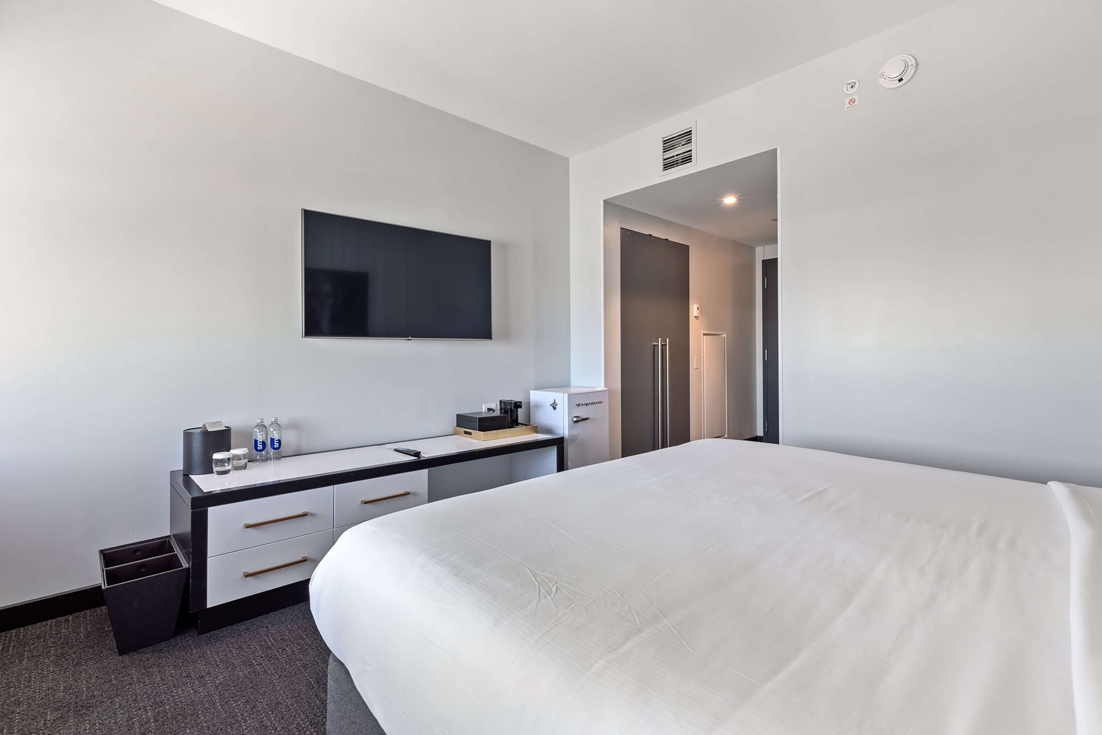 Quarto de hotel moderno com uma cama grande, uma escrivaninha com gavetas, uma televisão de tela plana na parede e uma lixeira pequena.