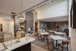 Uma sala de jantar moderna com mesas e cadeiras, um dispensador de suco e uma estante de livros ao fundo.