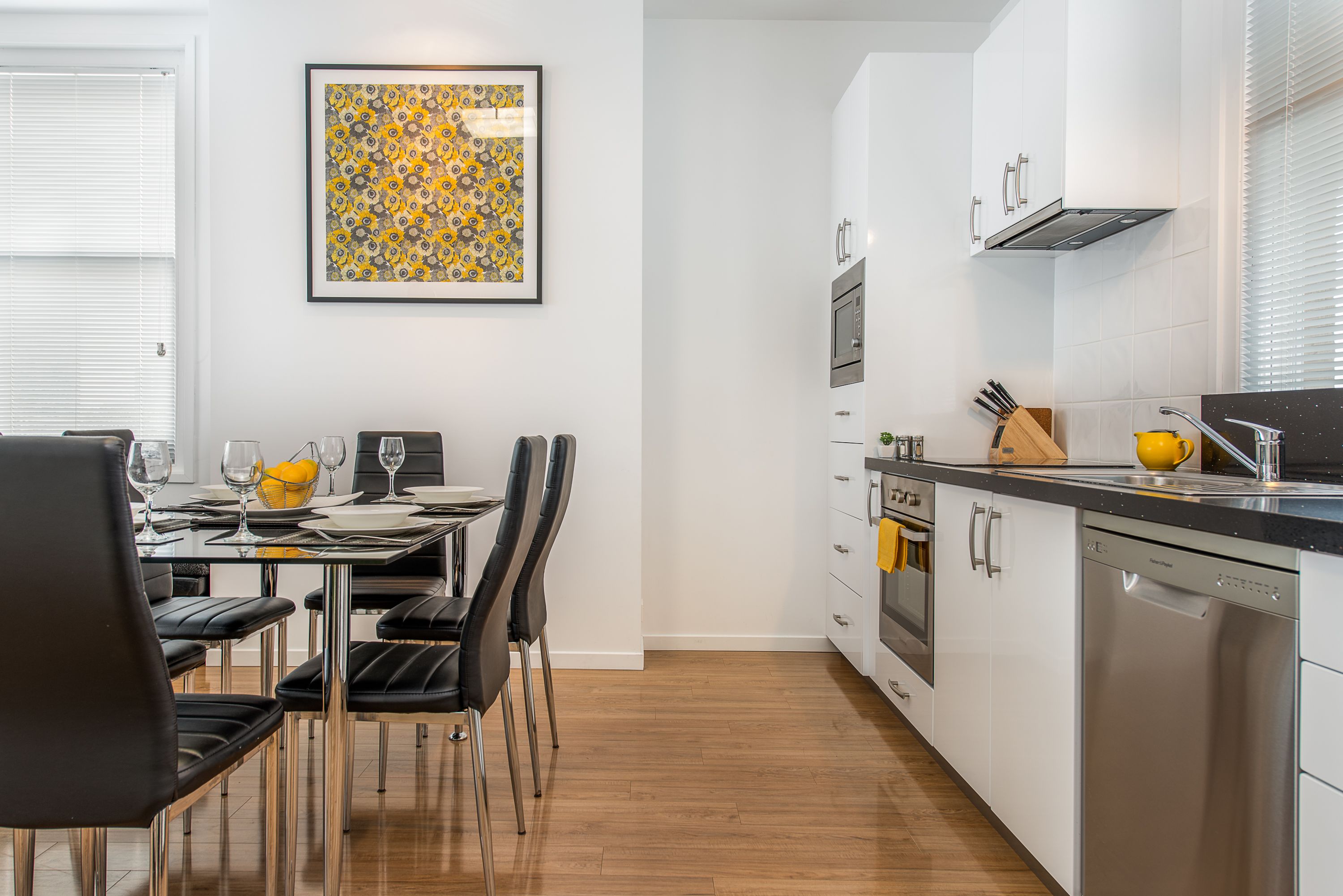 Uma cozinha moderna com armários brancos, eletrodomésticos de aço inoxidável e uma área de jantar com cadeiras pretas e uma mesa de vidro.