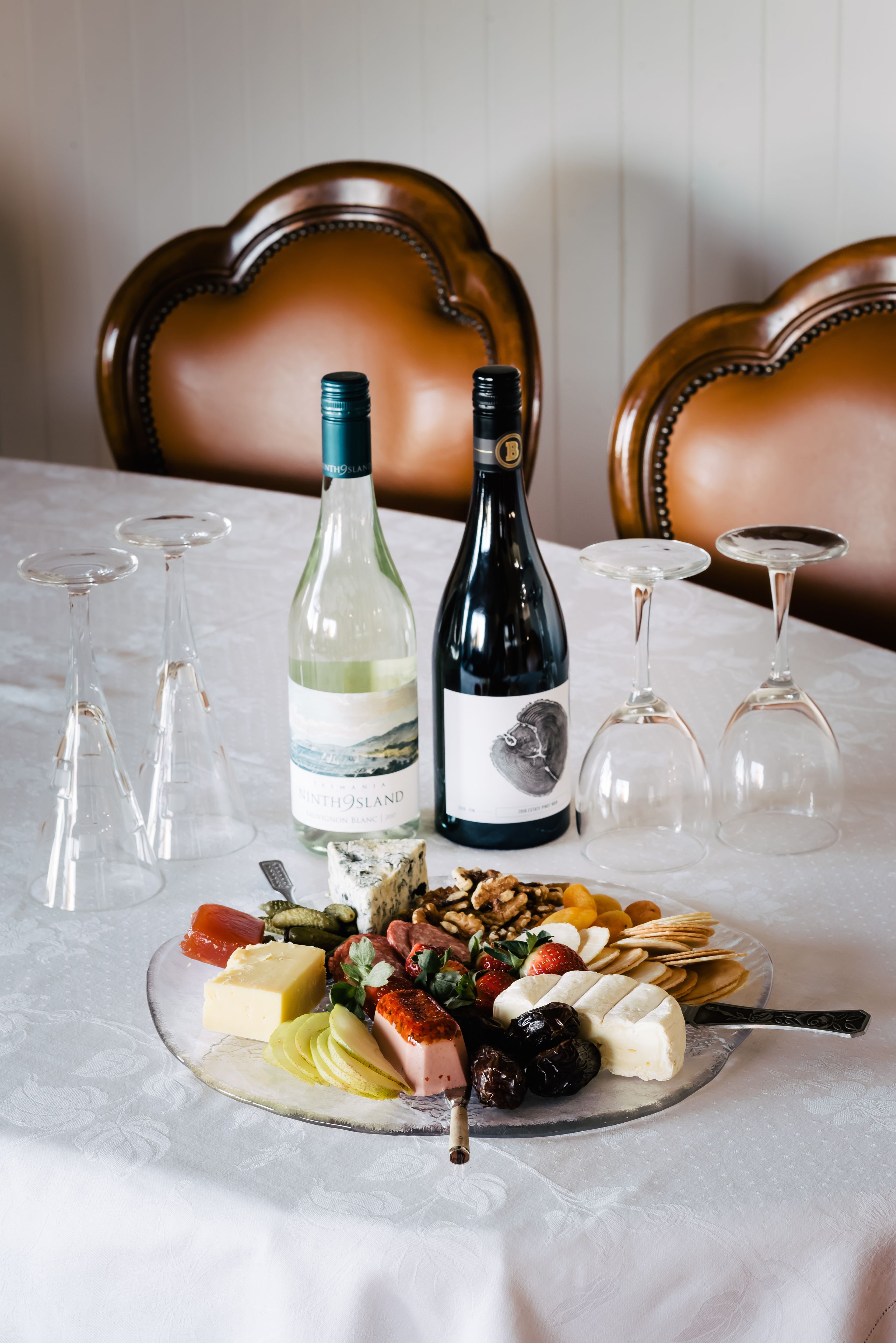 Uma mesa posta com um tábua de queijos, garrafas de vinho e copos sobre um lençol de mesa branco.