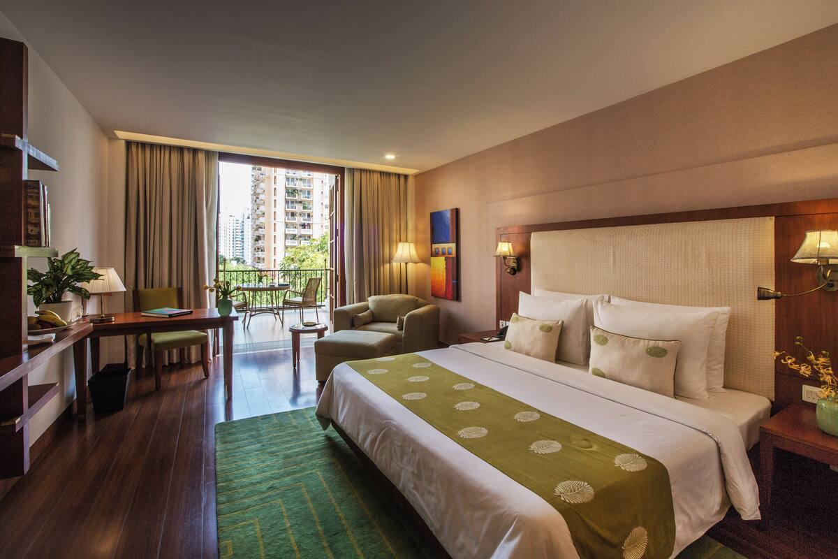 Quarto de hotel com uma cama grande, uma escrivaninha, uma cadeira, uma varanda com vista para edifícios e uma janela com cortinas.