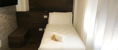 Uma cama pequena e bem arrumada com roupa de cama branca e uma toalha enrolada, colocada num canto com uma parede de madeira escura e um pequeno degrau.