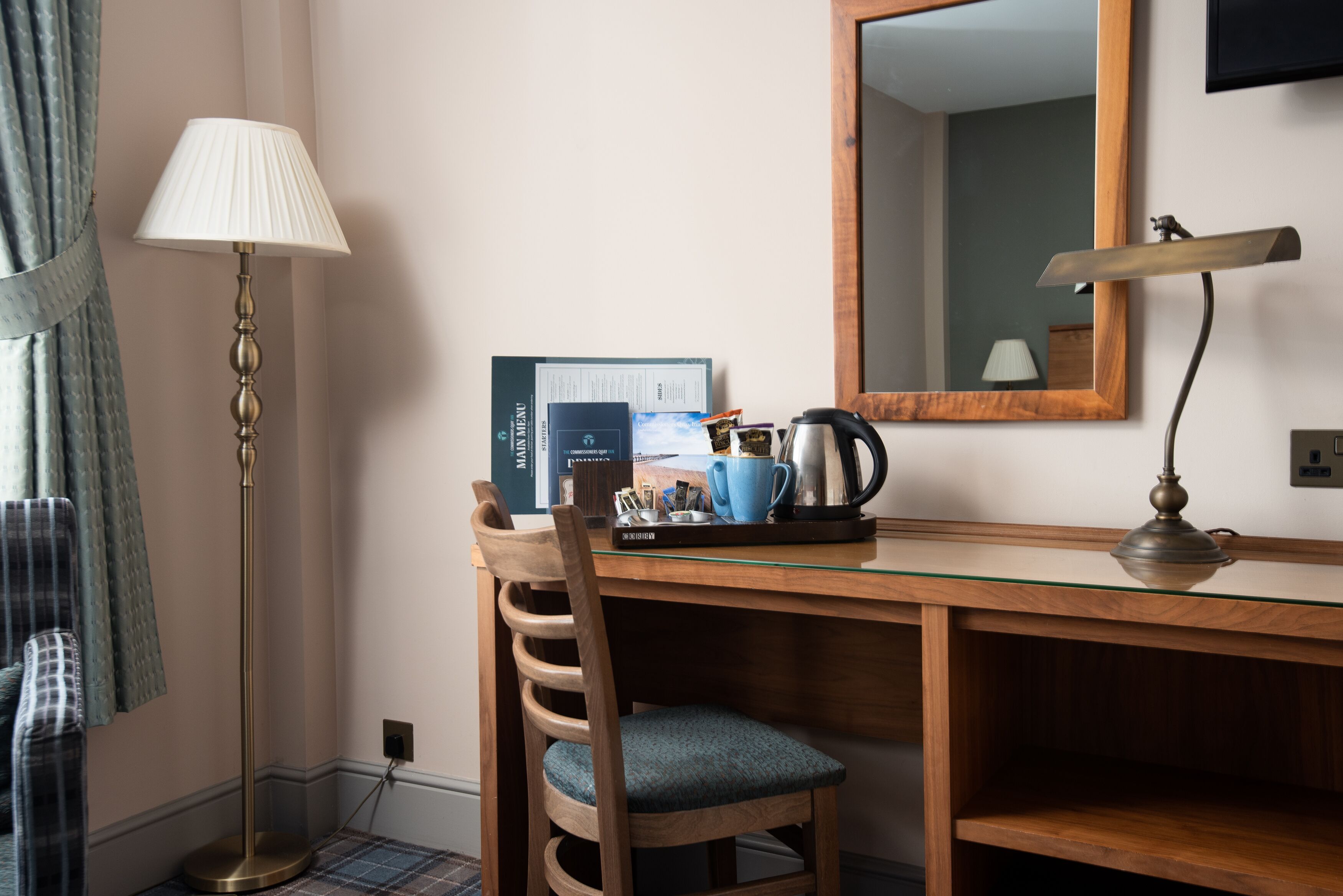Quarto de hotel com uma escrivaninha de madeira, uma cadeira, uma luminária, um espelho e uma janela com cortinas.