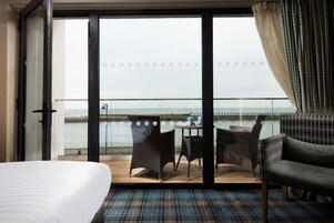 Quarto de hotel com uma grande porta de correr de vidro que dá para uma varanda com móveis de vime e vista para o mar.