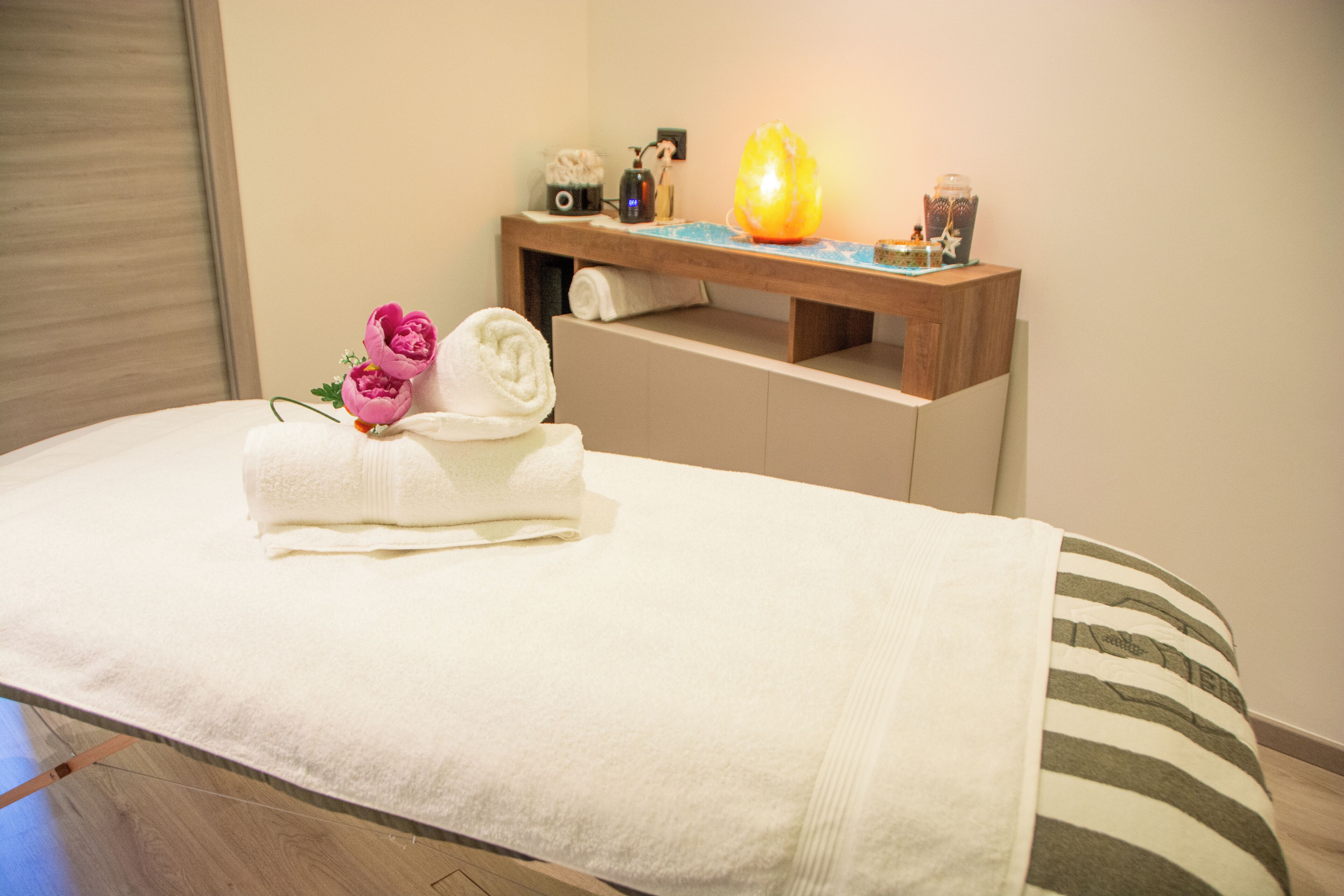 Cama bem arrumada com toalhas brancas e uma flor cor-de-rosa, prateleira de madeira com abajur e pequeno objeto decorativo.
