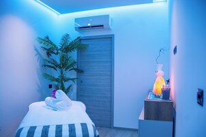 Quarto de hotel moderno com cama, planta em vaso, aparelho de ar condicionado e mesa de cabeceira com luminária.