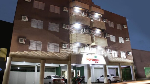 Um hotel com vários andares e uma placa com a inscrição "Portucali Hotel". Possui várias varandas, aparelhos de ar condicionado e um estacionamento com vários carros.