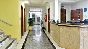 Um saguão de hotel com balcão de recepção, extintor de incêndio, planta em vaso e escada.