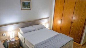 Uma cama bem arrumada com lençóis brancos, uma mesa de cabeceira com abajur, uma gavetinha e um guarda-roupa de madeira.