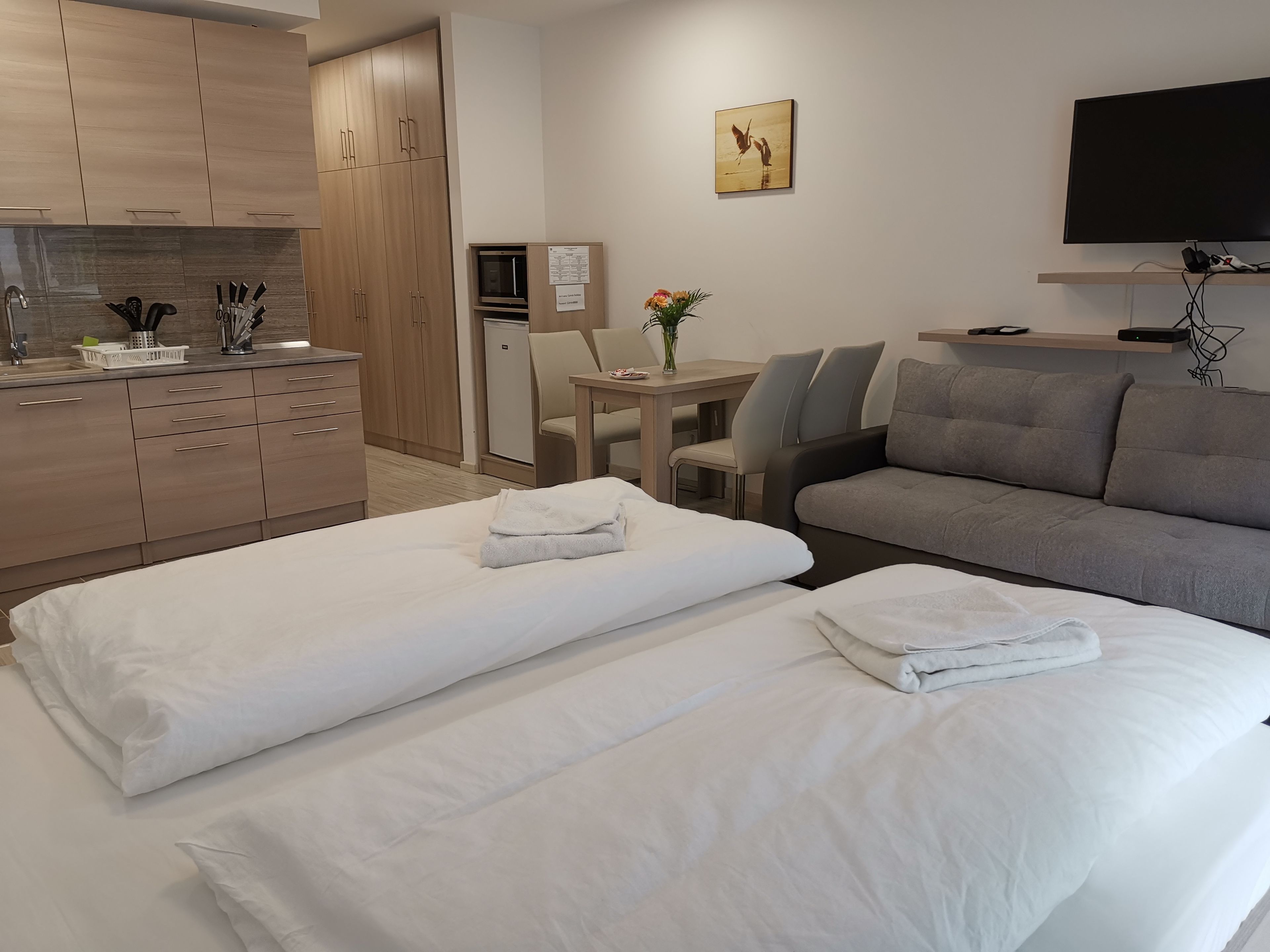 Quarto de hotel moderno com cozinha compacta, espaço de refeições, sofá cinza e duas camas com roupa de cama branca.