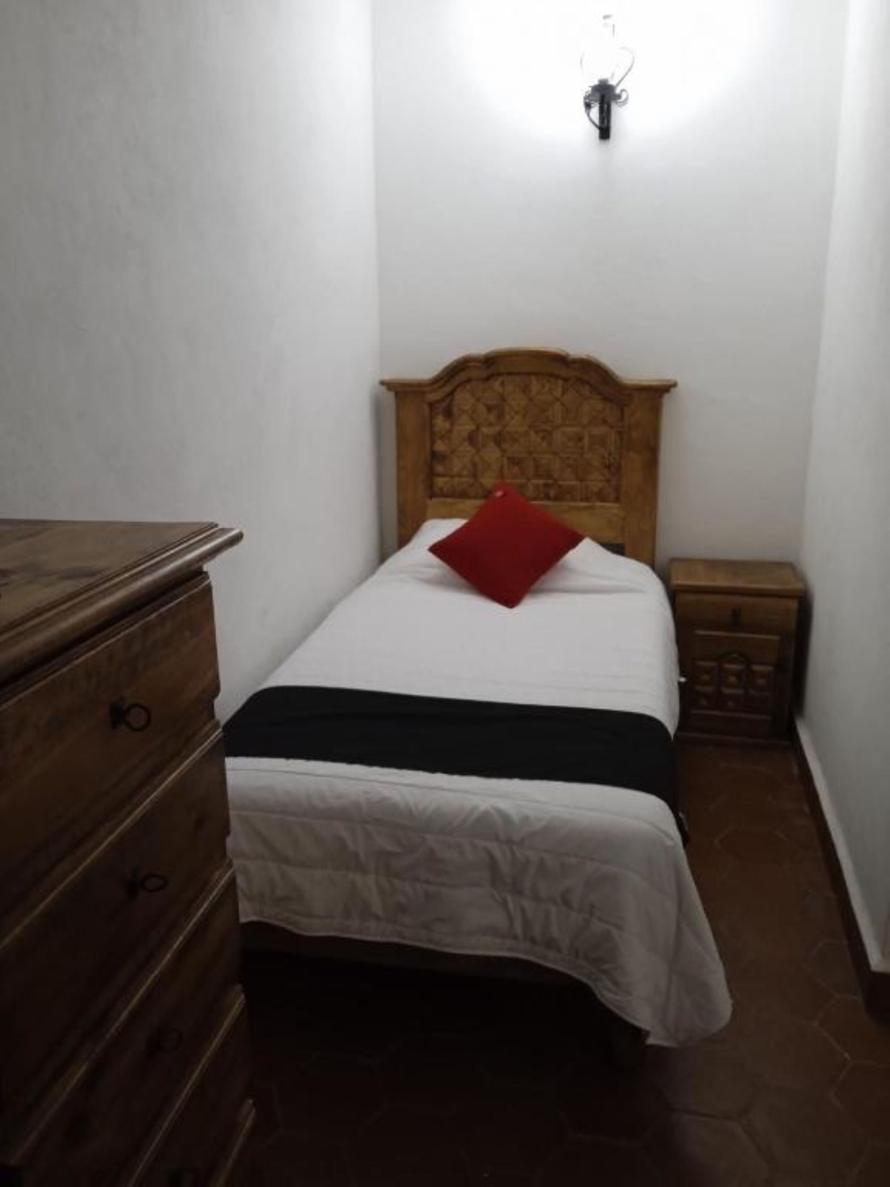 Quarto pequeno e bem arrumado, com cabeceira de madeira, uma cama de solteiro com travesseiro vermelho, cômoda de madeira e mesa de cabeceira.