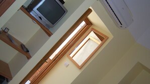 Um quarto com televisão na parede, ar condicionado, janela com moldura de madeira e uma pequena estátua em uma mesa.