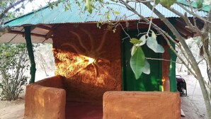 Uma cabana rústica com telhado de chapa metálica, uma porta verde e um pequeno alpendre com dois bancos de madeira.