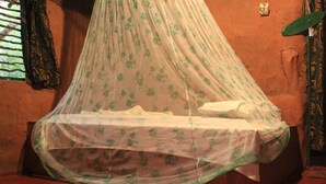 Uma cama com mosquiteiro, um banco de madeira, e uma parede com janela e porta.