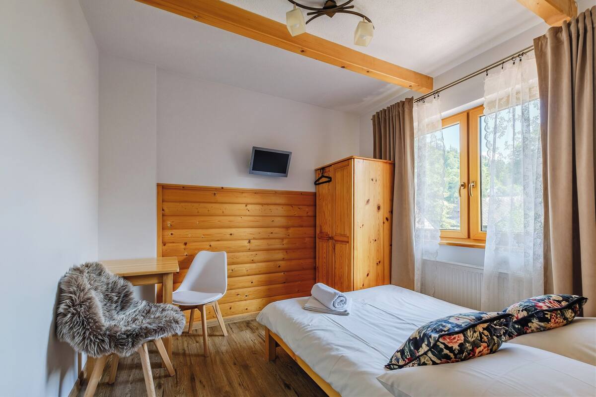 Um quarto com uma cama, uma cadeira, um guarda-roupa de madeira, uma televisão e uma janela com cortinas.