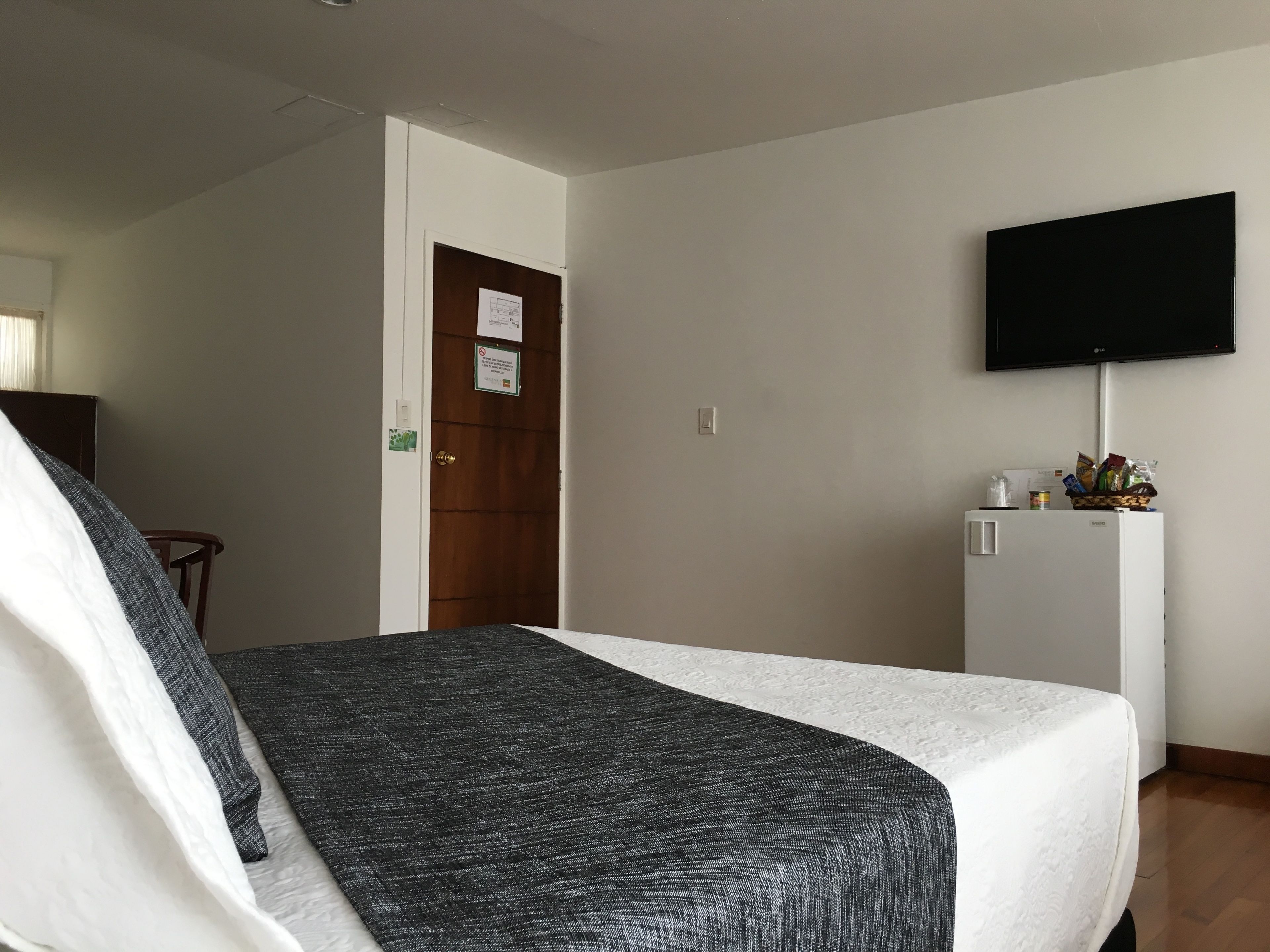 Quarto de hotel com uma cama, televisão na parede, armário branco e uma porta com uma placa.