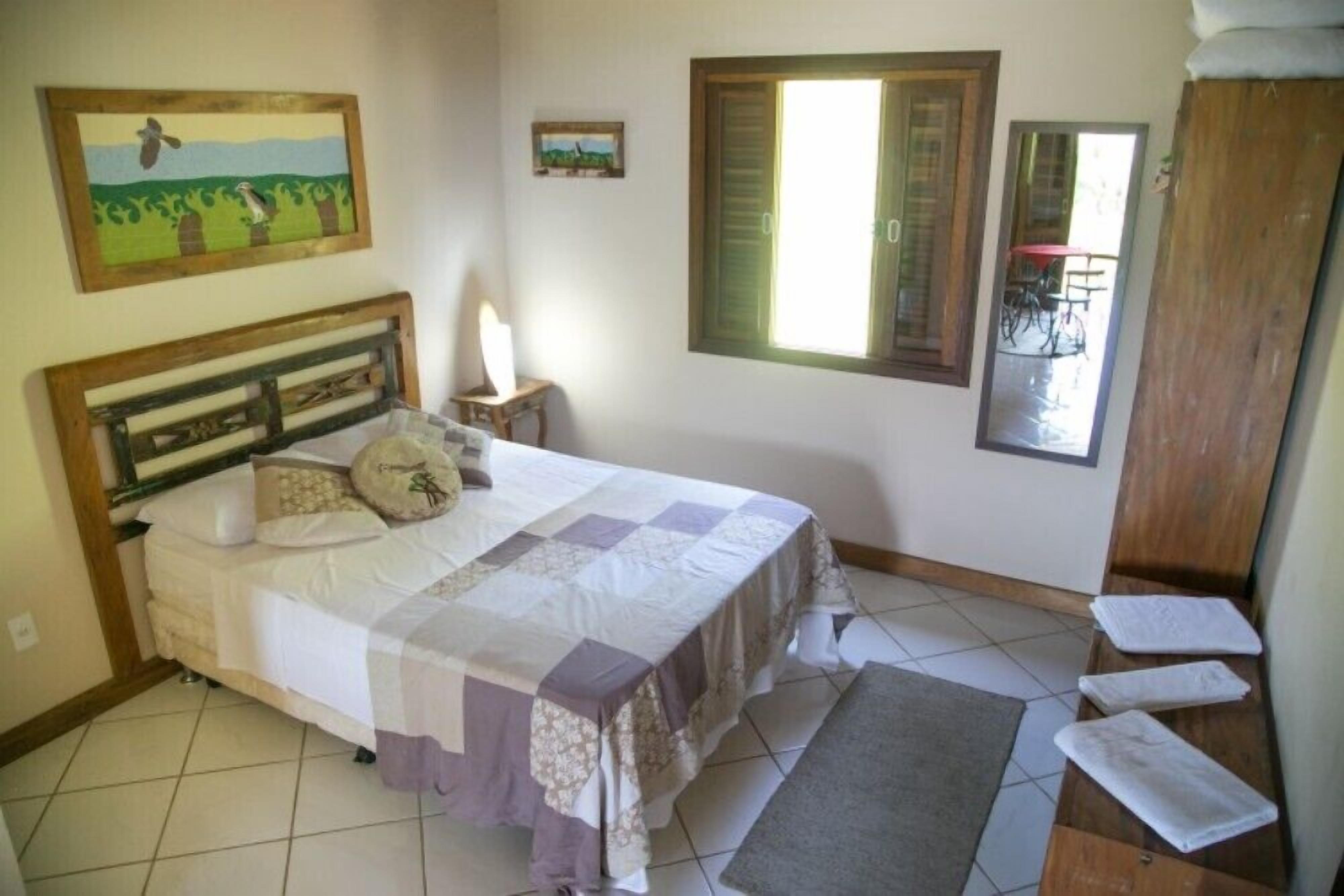 Quarto com cama, mesa de cabeceira, janela com venezianas e vista para o exterior.