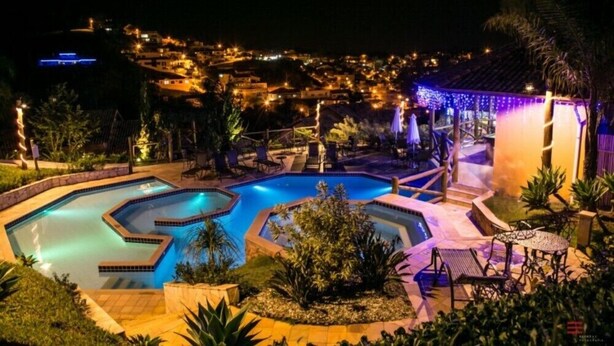 Vista noturna de um resort com uma ampla piscina com vários arcos, rodeada por áreas de estar ao ar livre e palmeiras.