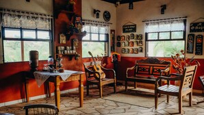 Quarto aconchegante com mesa de madeira, cadeiras e um violão. Janelas com cortinas de renda e vista para o verde lá fora.