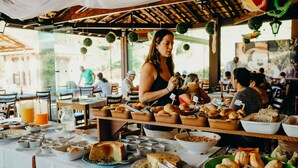 Disposição de buffet com diversos alimentos e uma mulher servindo bebidas.