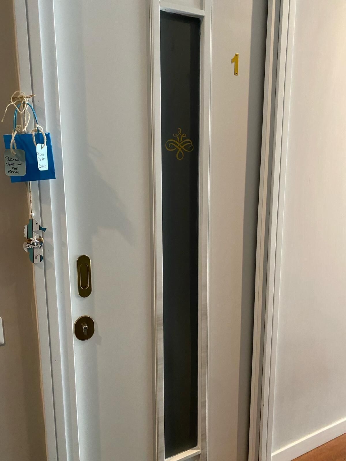 Uma porta branca com um painel de vidro, maçaneta de latão e uma pequena placa de latão com o número 1. Há duas etiquetas azuis com texto manuscrito na maçaneta.