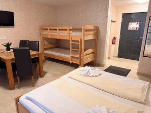 Uma cama beliche com escrivaninha e cadeiras, uma cama de solteiro com cobertor, e uma porta com placa.