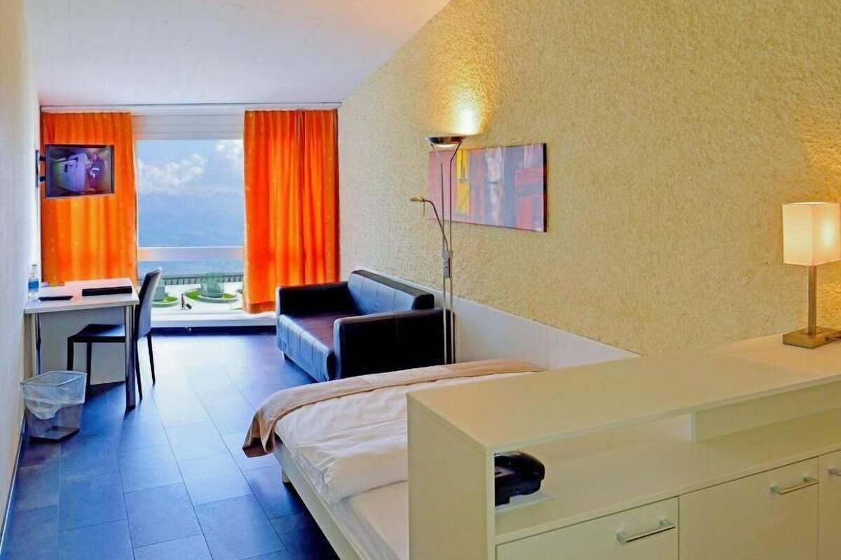 Quarto de hotel com uma cama, uma mesa com cadeira, um sofá, uma TV e uma janela com cortinas.
