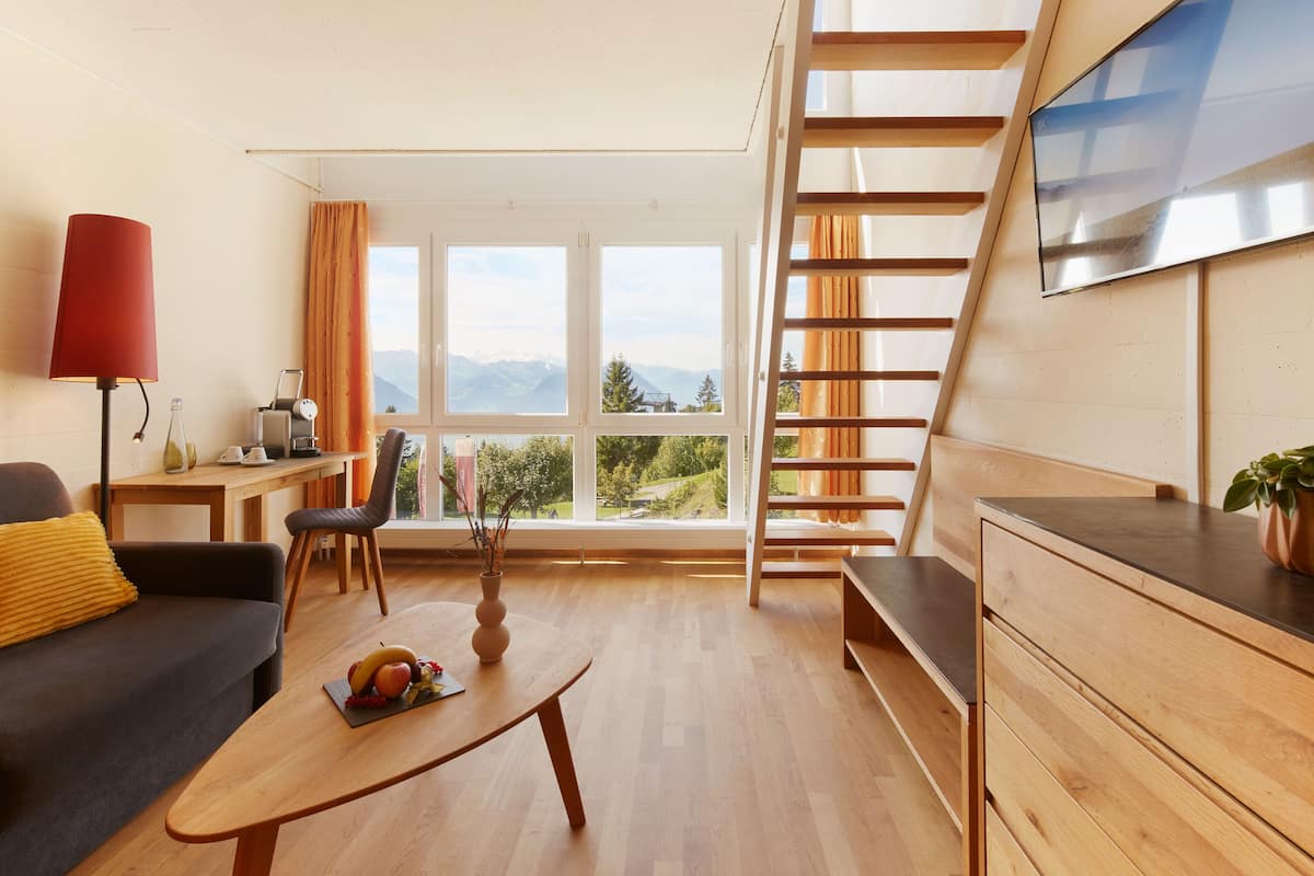 Uma sala de estar moderna com piso de madeira, um sofá, uma mesa de centro com frutas, uma escrivaninha com uma luminária, uma TV na parede e vista para montanhas através de janelas amplas.