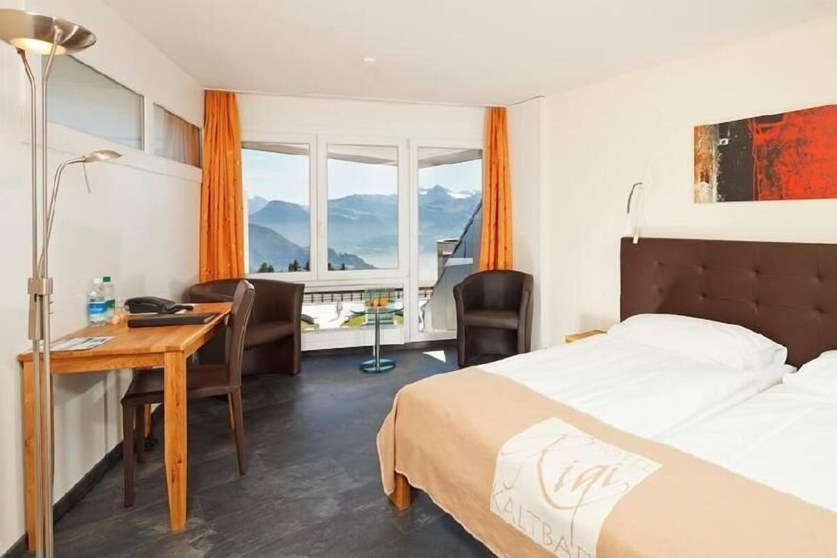 Quarto de hotel com cama, escrivaninha, cadeira e vista para montanhas.
