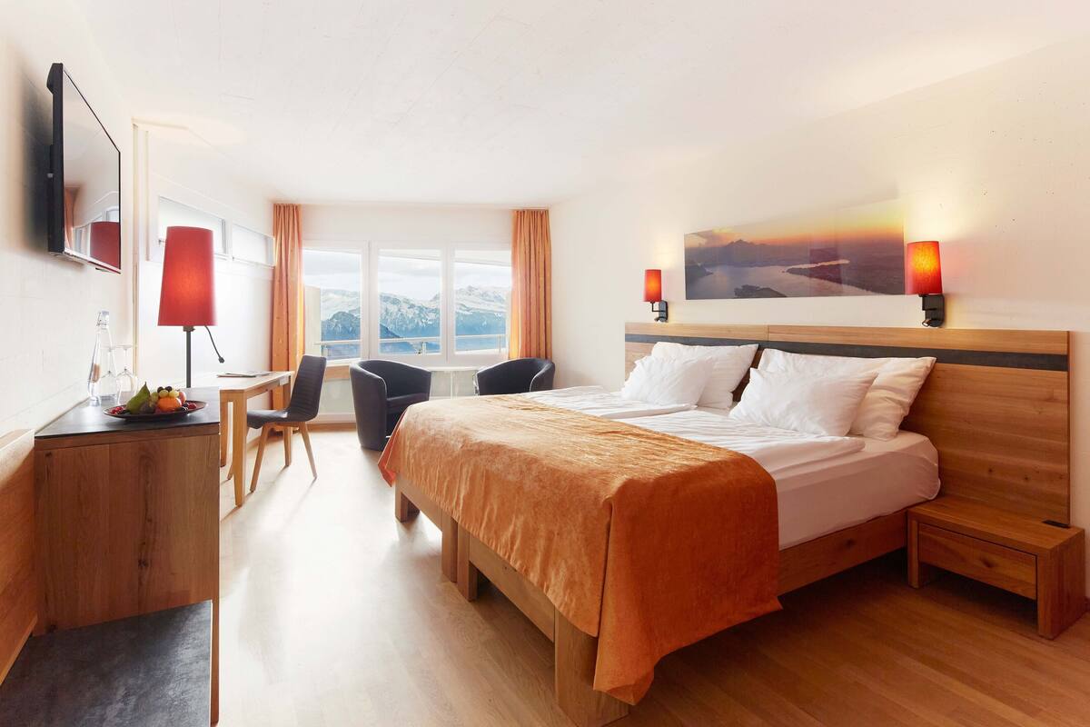 Quarto de hotel com cama de madeira, escrivaninha com cadeira, televisão e janela com vista para as montanhas.