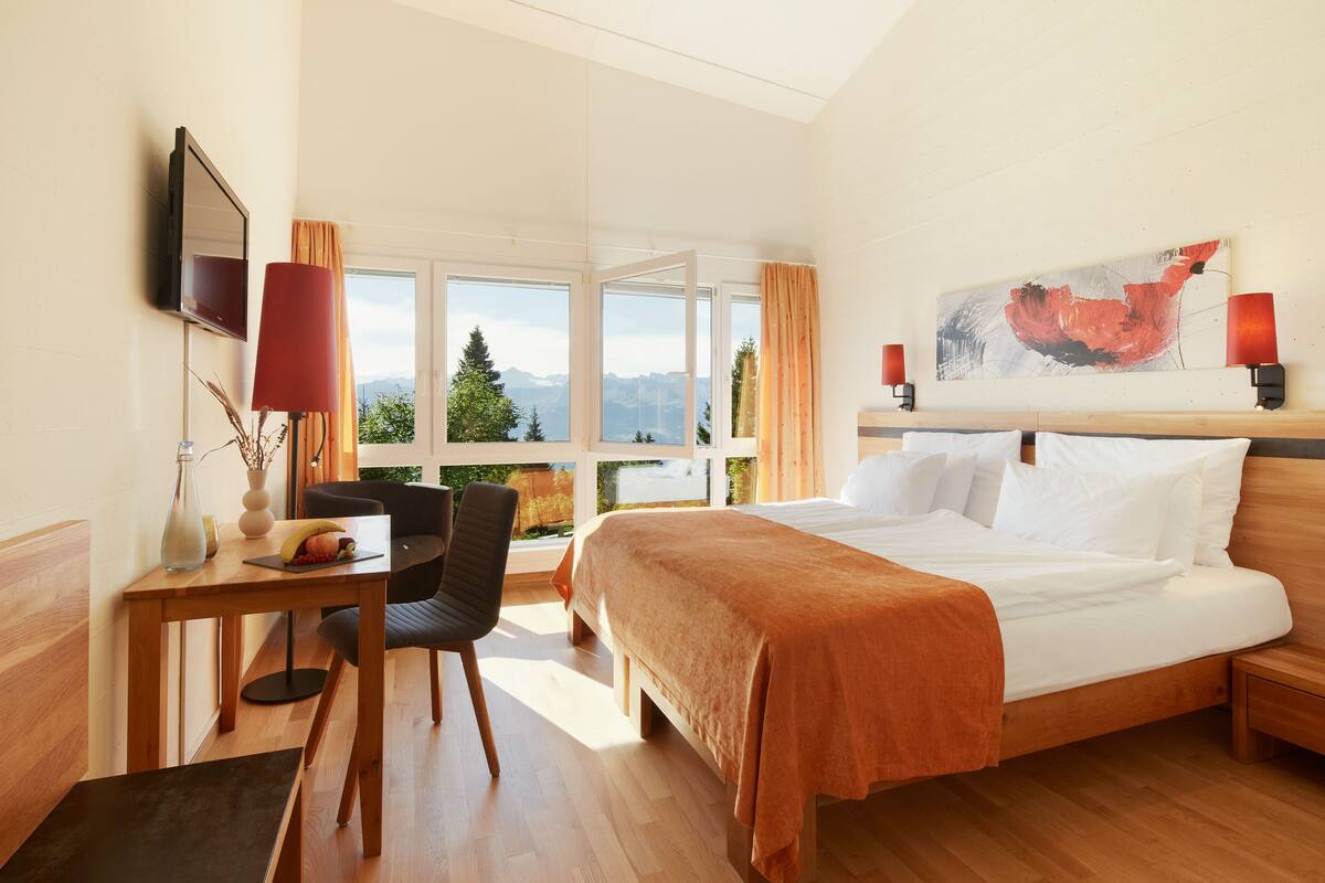 Quarto de hotel com cama, escrivaninha, cadeira, televisão e janela com vista para montanhas.