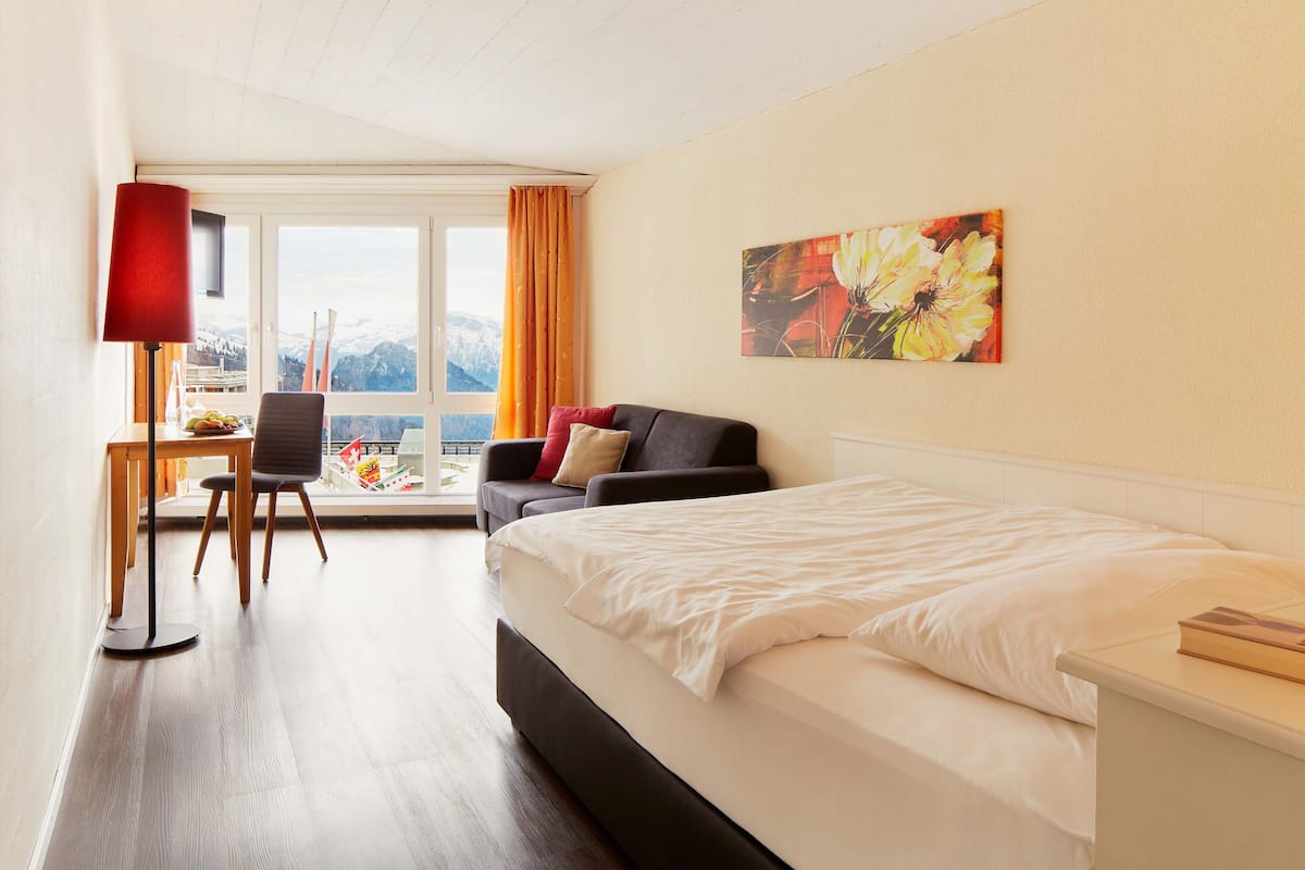 Quarto de hotel com uma cama, escrivaninha com cadeira, luminária vermelha e uma janela grande com vista para montanhas.