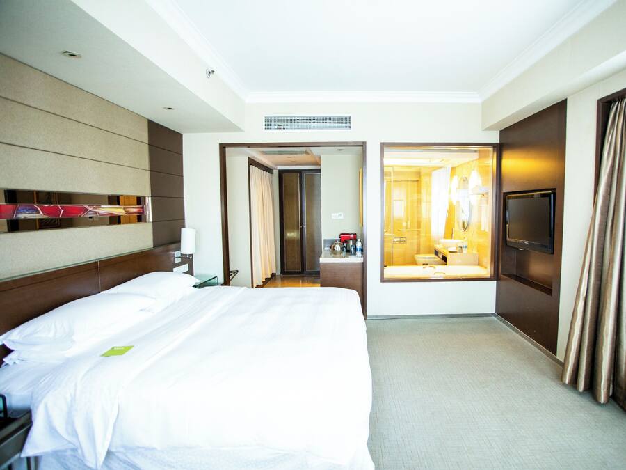Quarto de hotel com uma cama grande, televisão de tela plana, banheiro com espelho e pia, e vista para o saguão do hotel.
