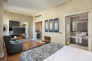 Quarto de hotel moderno com uma cama grande, área de estar com uma poltrona e uma mesinha, televisão e banheiro com pia e espelho.