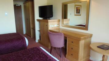 Quarto de hotel com duas camas, uma escrivaninha de madeira com cadeira, televisão, espelho e uma mesinha redonda com um livro.