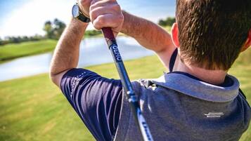 Uma pessoa segurando um taco de golfe, usando um relógio no pulso, em um campo de golfe com um corpo d'água ao fundo.
