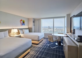 Quarto de hotel com duas camas, uma escrivaninha, uma cadeira e uma janela grande com vista para a cidade.