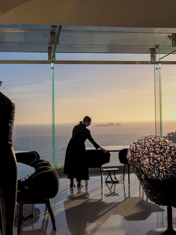 Interior moderno com guarda-corpo de vidro, uma pessoa sentada perto de uma mesa, e vista para o oceano.
