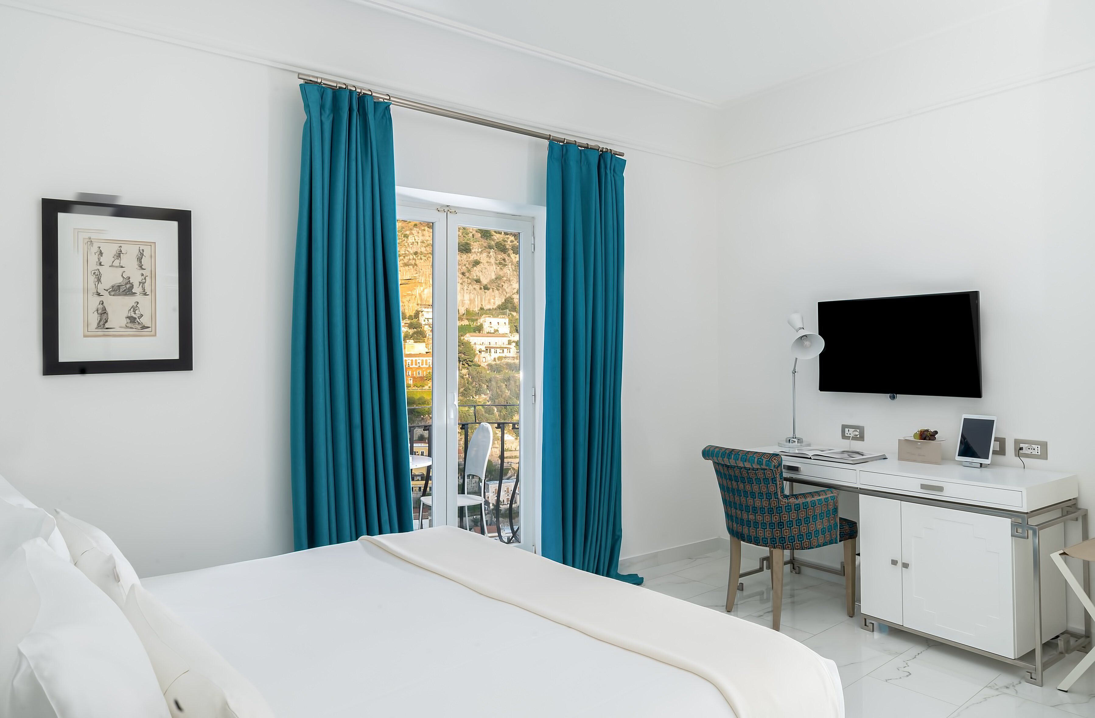 Quarto de hotel com cama, secretária com computador, televisão e vista para uma varanda com cortinas azuis.