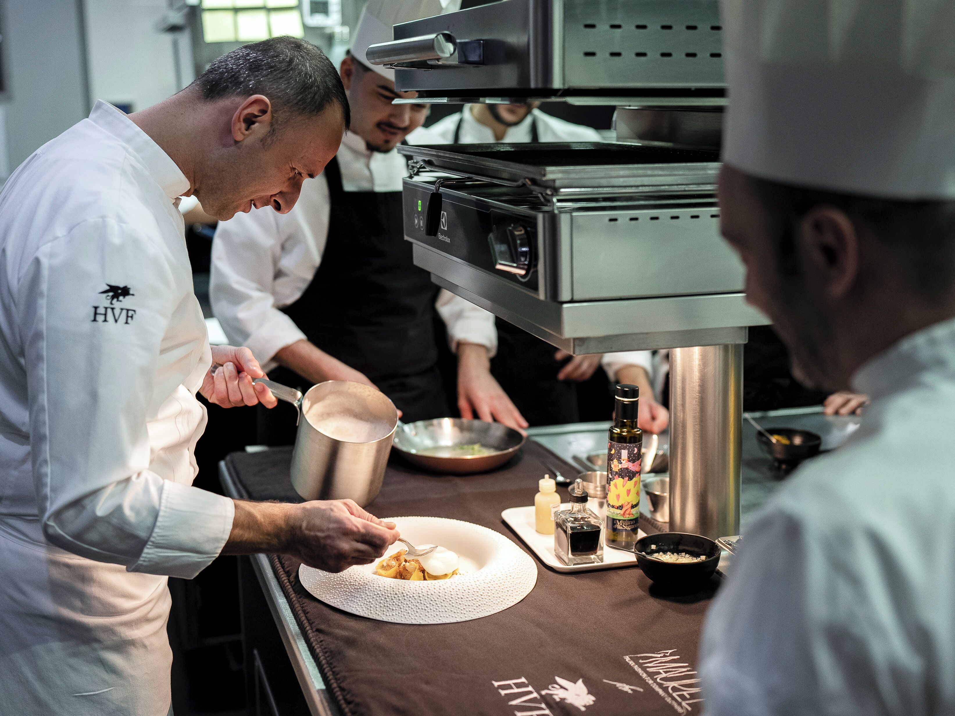 Um chefe de cozinha prepara alimentos em uma cozinha profissional, equipada com eletrodomésticos de aço inoxidável e um espaço de trabalho limpo e organizado.