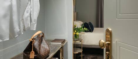 Quarto de hotel com uma cama, uma mesa de toucador com espelho, um roupão, uma bolsa e uma pequena mesa com um livro e flores.