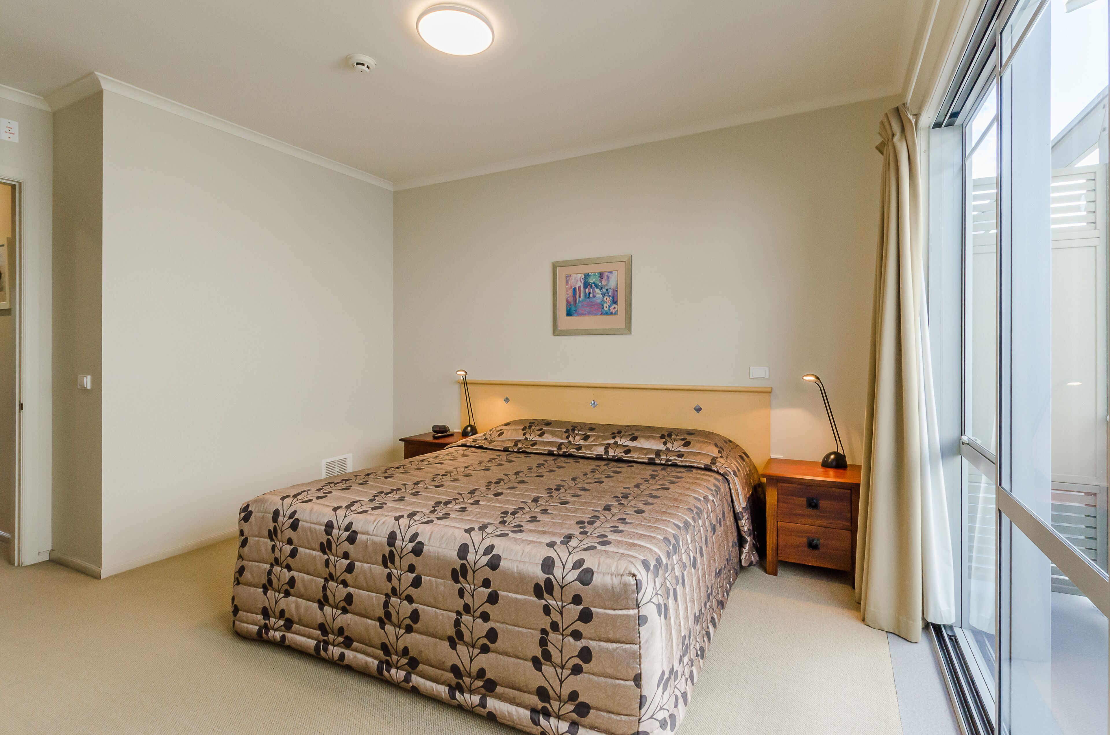 Quarto com uma cama grande, mesas de cabeceira, um criado-mudo, um quadro emoldurado na parede e uma janela com cortinas.