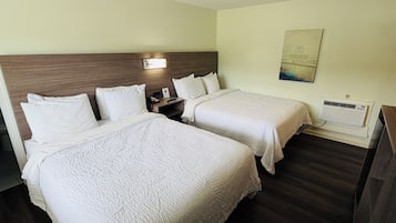 Quarto de hotel com duas camas, uma mesa de cabeceira, ar condicionado fixado na parede e uma citação emoldurada.