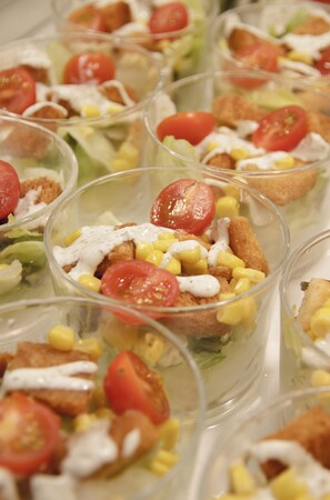 Close-up de vários copos de vidro transparentes repletos de salada com tomate, milho e croutons, coberta por um molho branco.