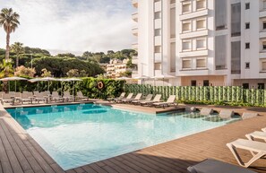 Uma área de piscina com espreguiçadeiras e guarda-sóis, cercada por um edifício branco com varandas.