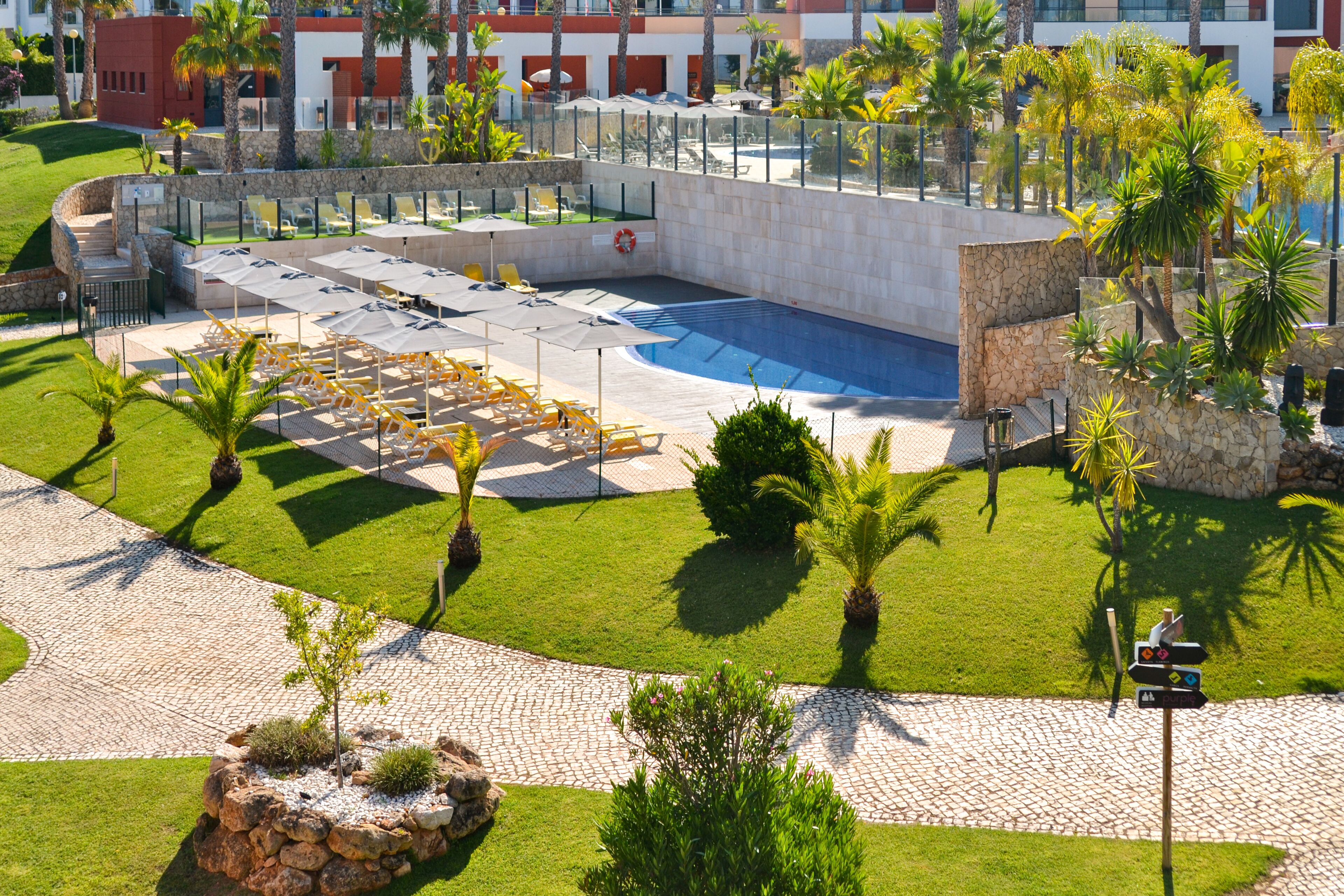Uma área de piscina com espreguiçadeiras e guarda-sóis, cercada por palmeiras e um muro de pedra.