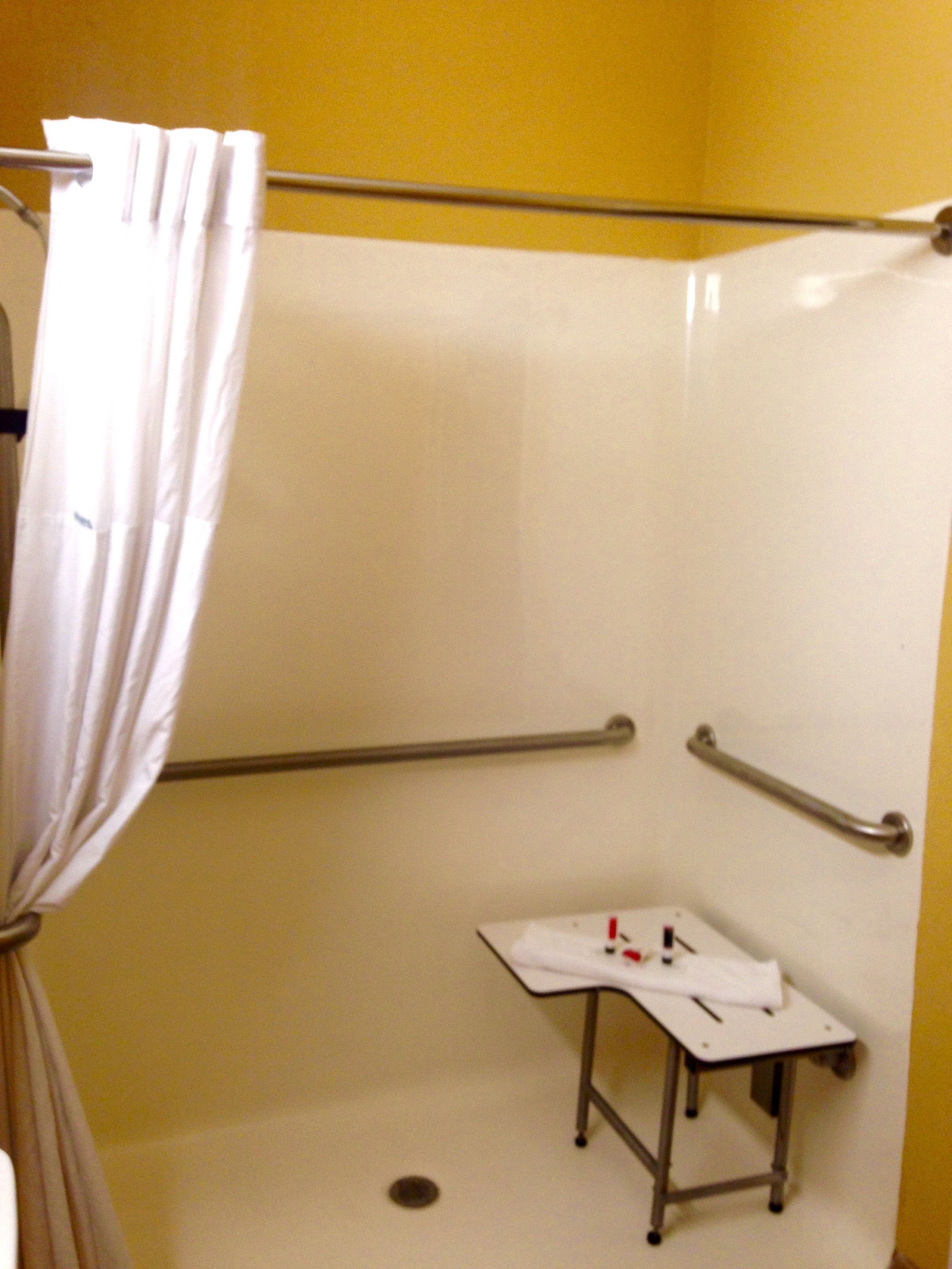 Área de duche com cortina branca, barras de apoio e uma pequena mesa com produtos de higiene pessoal.